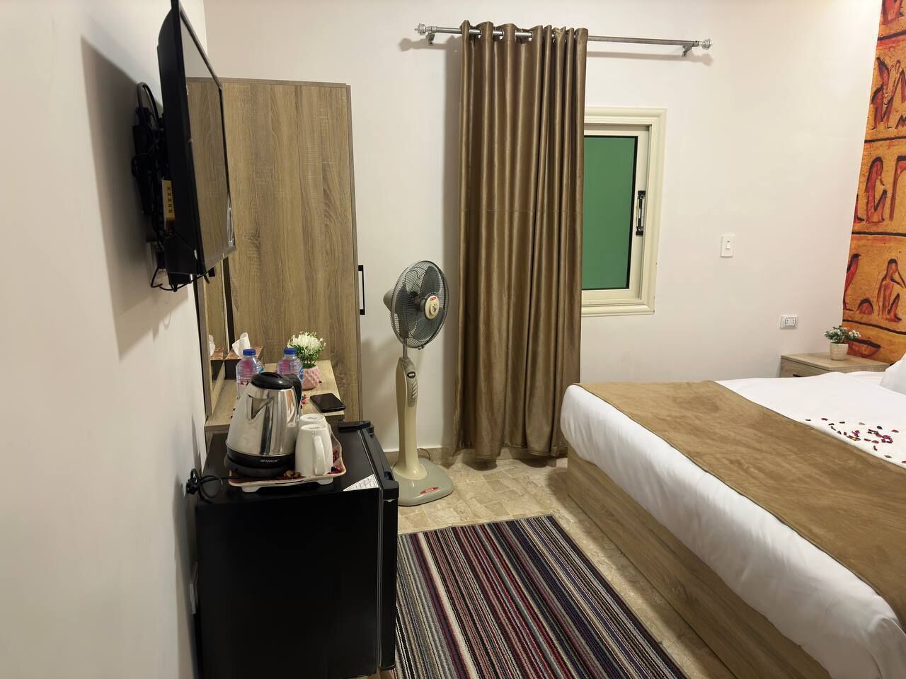 Quarto de hotel com cama, escrivaninha com chaleira, ventoinha e cortina.