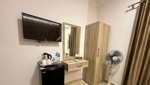 Quarto de hotel com televisão de ecrã plano, minibar, guarda-roupa de madeira, ventoinha e tapete riscado.