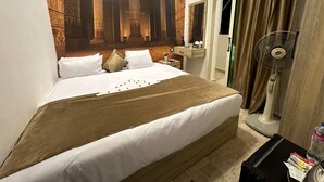 Quarto de hotel com uma cama grande, cabeceira de madeira, mesa de cabeceira, ventilador e espelho.