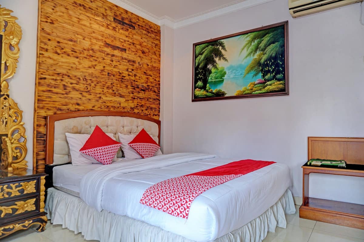 Quarto com cabeceira de madeira, cama grande com roupa de cama branca e detalhes vermelhos, pintura de paisagem tropical e mesa de cabeceira de madeira com gavetas.