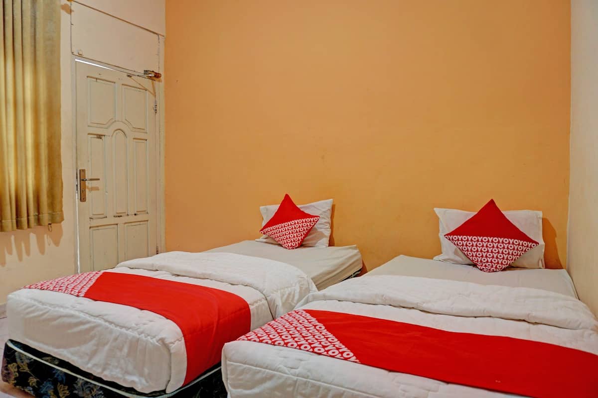 Quarto com duas camas, cada uma com roupa de cama branca e detalhes vermelhos, e uma porta fechada com interruptor de luz acima.