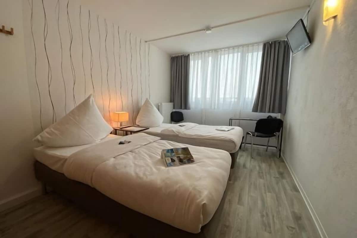 Quarto de hotel com duas camas, uma escrivaninha, uma cadeira, uma televisão e uma janela com cortinas.