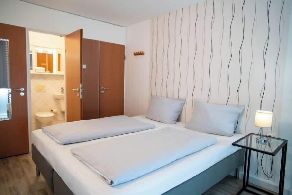 Uma cama de casal com duas almofadas, uma mesa de cabeceira com abajur, um guarda-roupa de madeira e um banheiro visível por uma porta aberta.