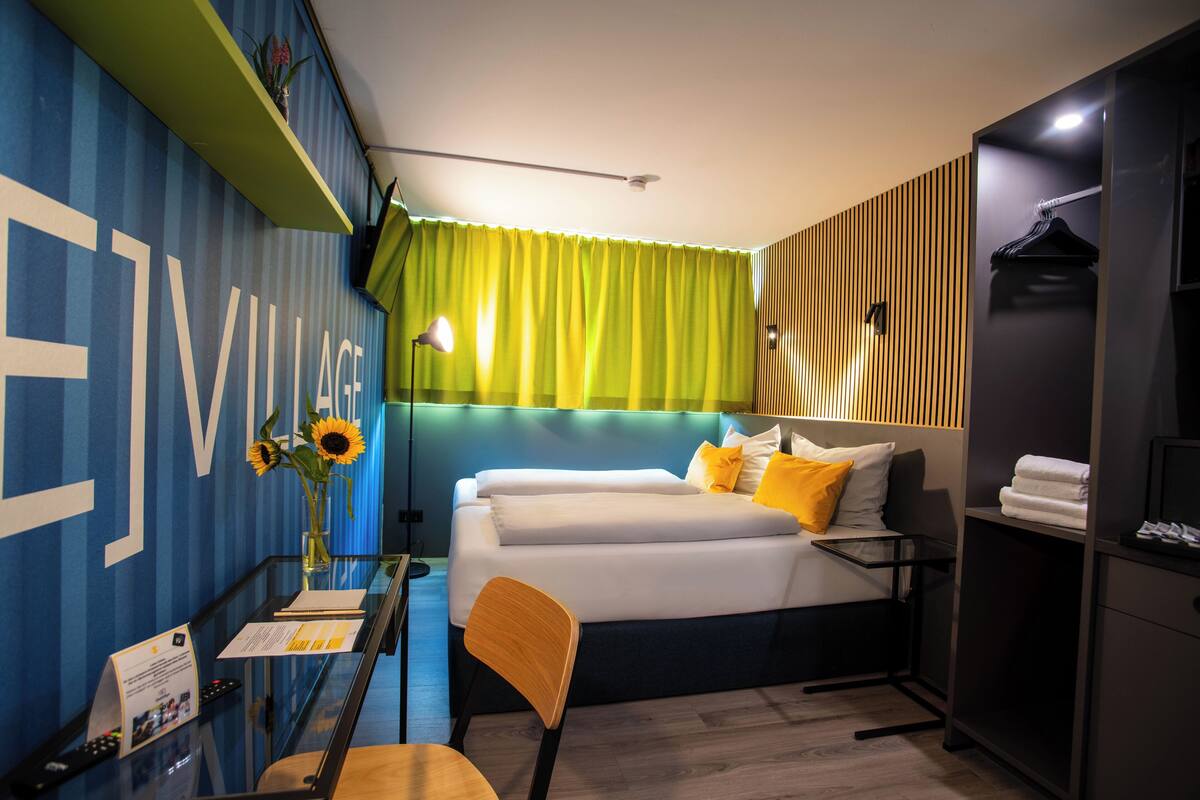 Quarto de hotel moderno com uma cama grande, uma escrivaninha com cadeira, uma mesa de vidro e um guarda-roupa.