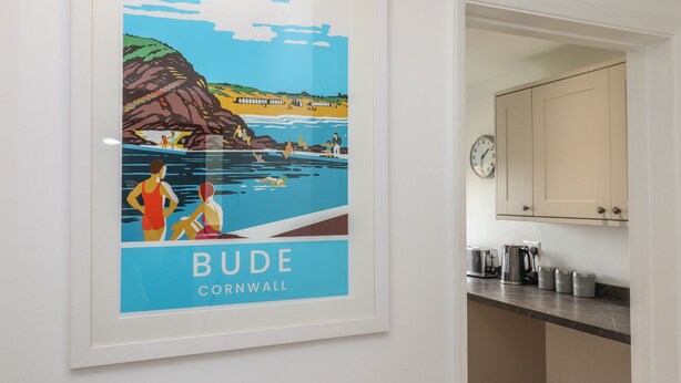 Um quadro com um cartaz de uma praia, exibindo o texto "Bude Cornwall", pendurado em uma parede branca.