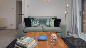 Uma sala de estar com um sofá cinzento, uma mesa de centro com livros e um vaso de vidro, e um candeeiro fixado na parede.