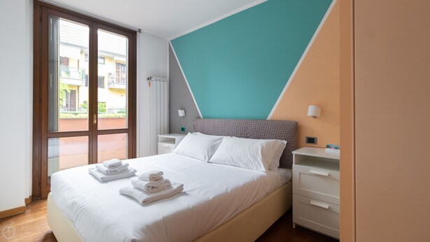 Quarto moderno com cama, mesas de cabeceira brancas, parede em tom de verde-água e vista para o exterior.
