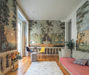 Um quarto com um grande mural, piso de madeira, sofá com almofadas estampadas, uma pequena mesa com cadeiras e um vaso com flores.