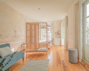 Quarto com piso de madeira, sofá azul, estrutura de cama de madeira, mesa pequena e um banquinho cinza.