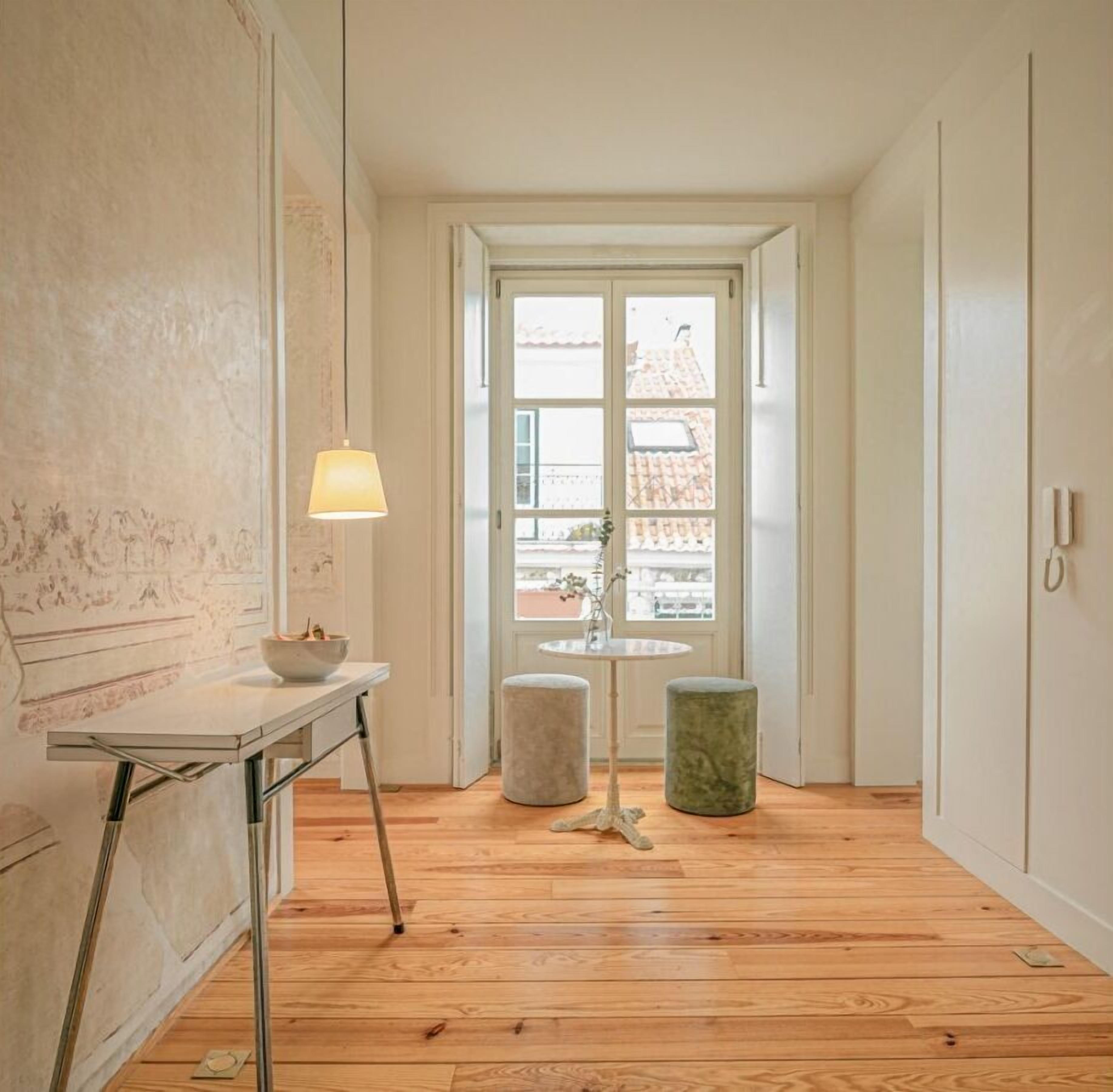 Um corredor estreito com piso de madeira, uma porta branca, uma mesinha com abajur e dois pufes.