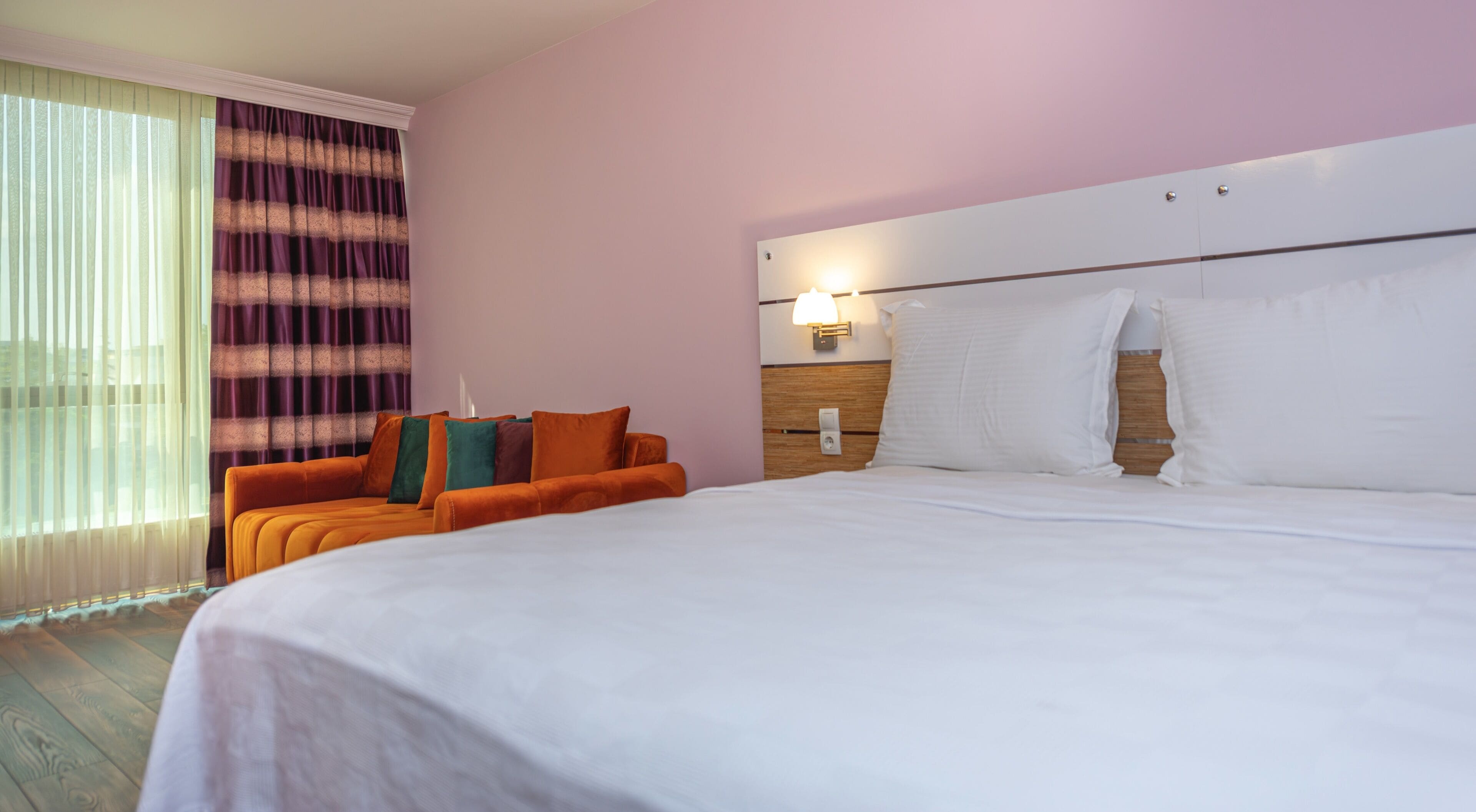 Quarto de hotel com uma cama grande, um sofá com almofadas, uma mesa de cabeceira com abajur e uma janela com cortinas.