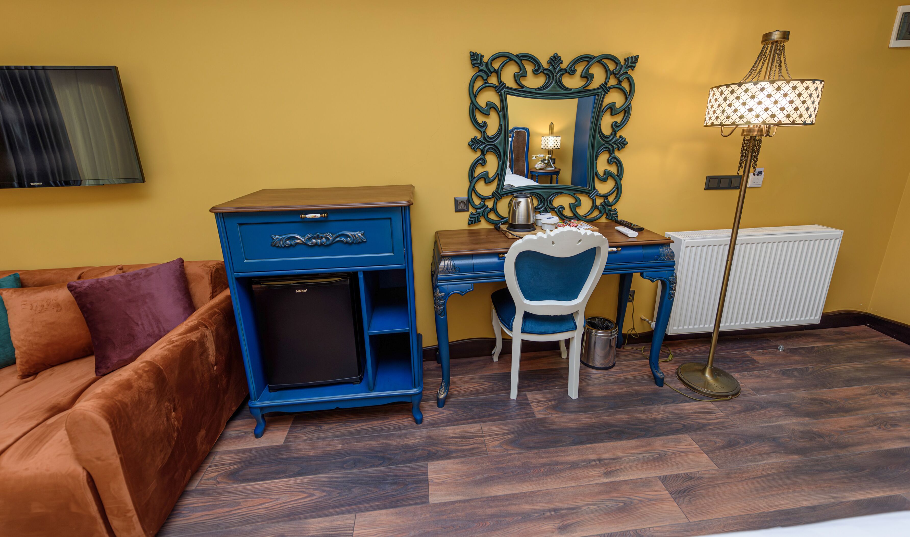 Quarto compacto com uma escrivaninha azul, uma cadeira branca, um pequeno frigorífico, um radiador e uma TV fixada na parede.