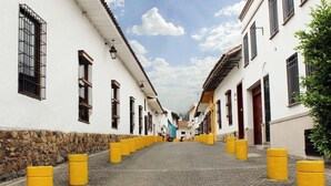 Uma rua de calçada estreita, ladeada por edifícios brancos com grades nas janelas e mourões amarelos.
