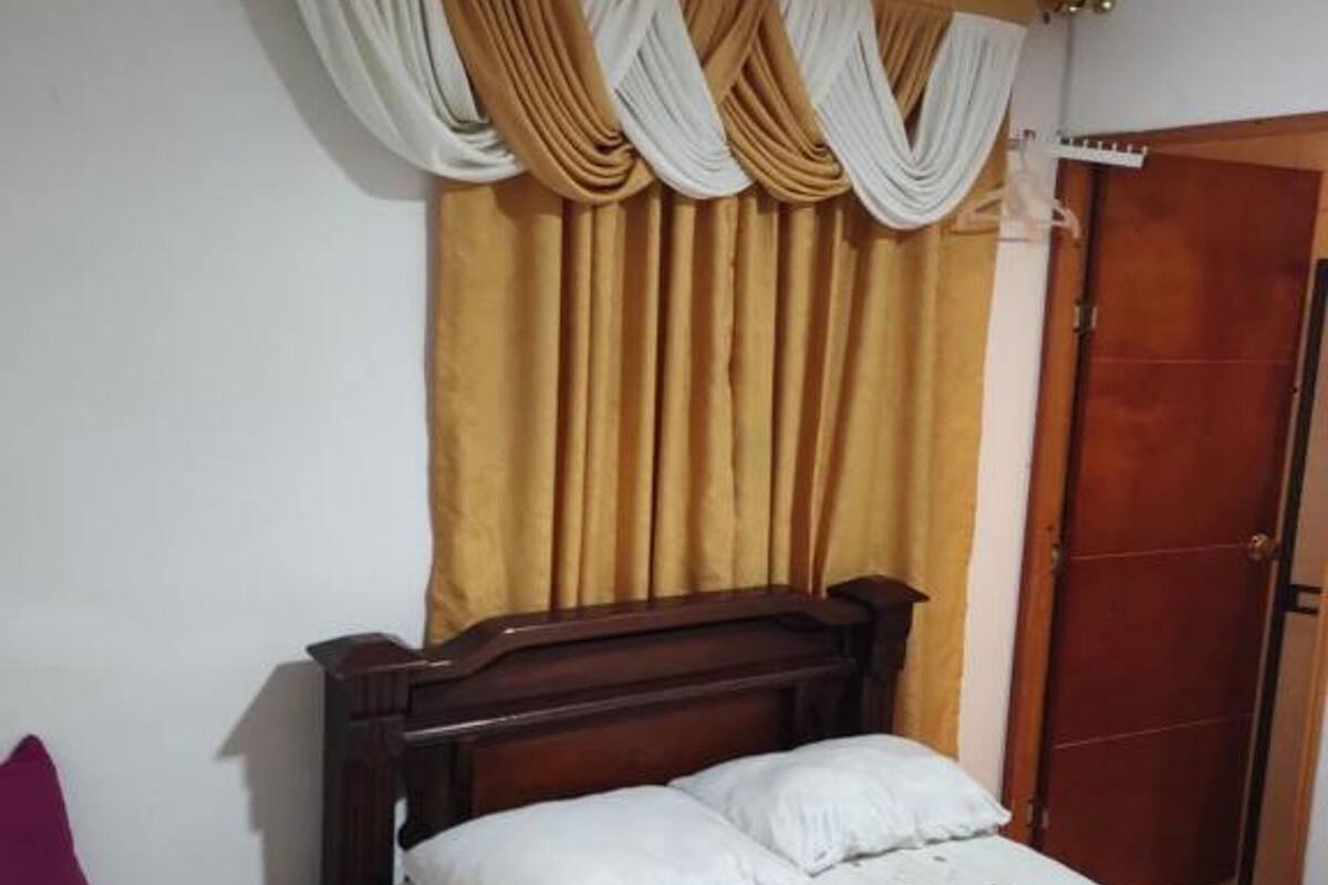 Um quarto com cama de madeira, roupa de cama branca, um guarda-roupa de madeira e uma cortina amarela.