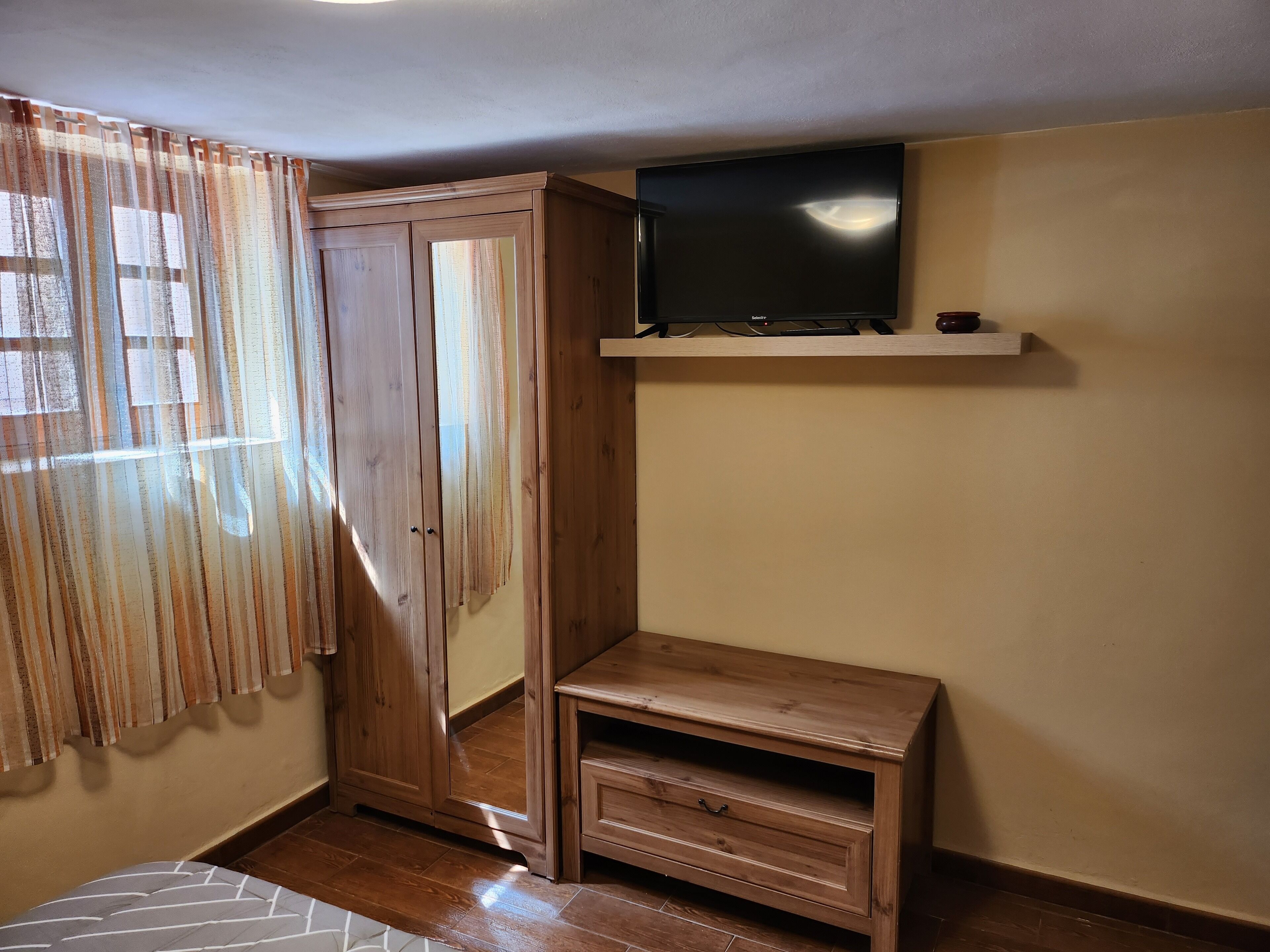 Quarto com guarda-roupa, televisão presa à parede e uma pequena mesa de cabeceira de madeira.