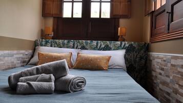 Cama bem arrumada com colcha azul, duas almofadas estampadas com flores e toalhas enroladas.
