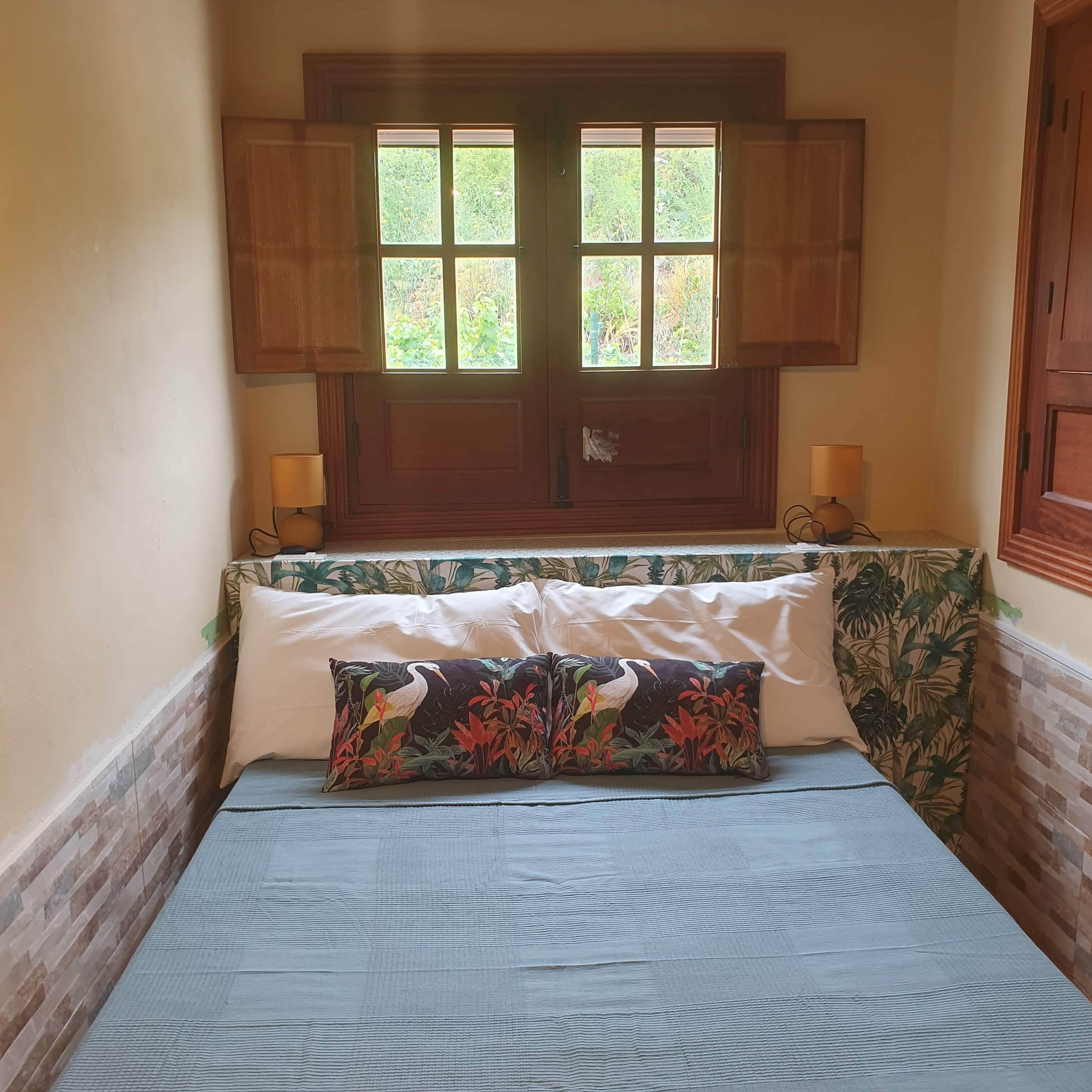 Quarto com cama, janela com vista para vegetação, armários de madeira e duas luminárias fixadas na parede.