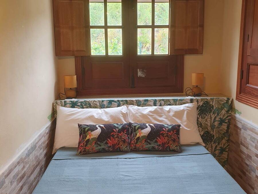 Quarto com cama, janela com vista para vegetação, armários de madeira e duas luminárias fixadas na parede.