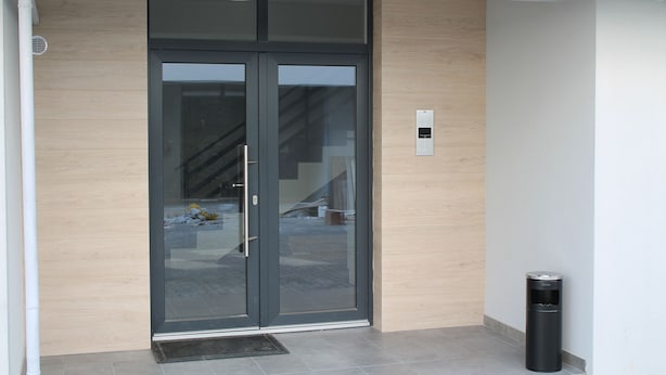 Entrada de um edifício moderno com porta de vidro, lixeira de metal e um pequeno degrau até a porta.