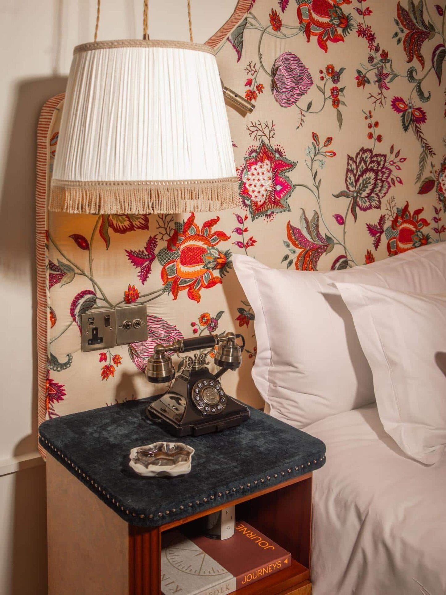 Um quarto com uma cama, uma mesa de cabeceira com abajur, um telefone giratório antigo e um livro intitulado 'Journeys'.