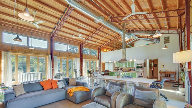 Uma sala de estar espaçosa, com um sofá grande, pufes e poltronas, uma cozinha ao fundo e um teto alto com vigas aparentes.