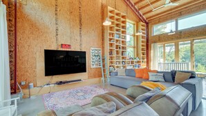 Uma sala de estar ampla com uma grande TV de tela plana na parede, um sofá cinza, uma estante de madeira e uma janela com vista para o exterior.