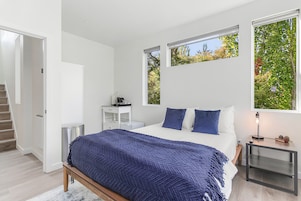 Quarto com cama, mesa de cabeceira, cômoda e janela com vista para árvores.
