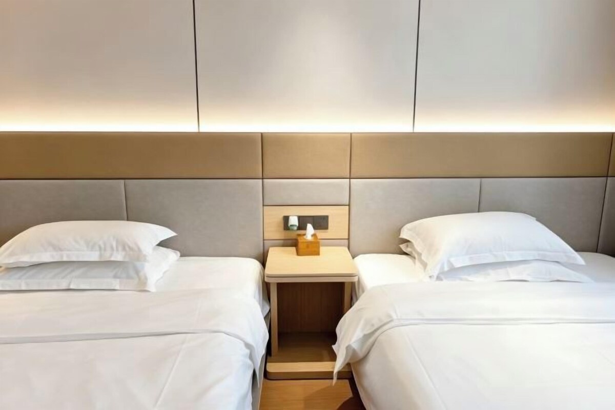 Quarto de hotel com duas camas, uma mesa de cabeceira e luminária fixa na parede.
