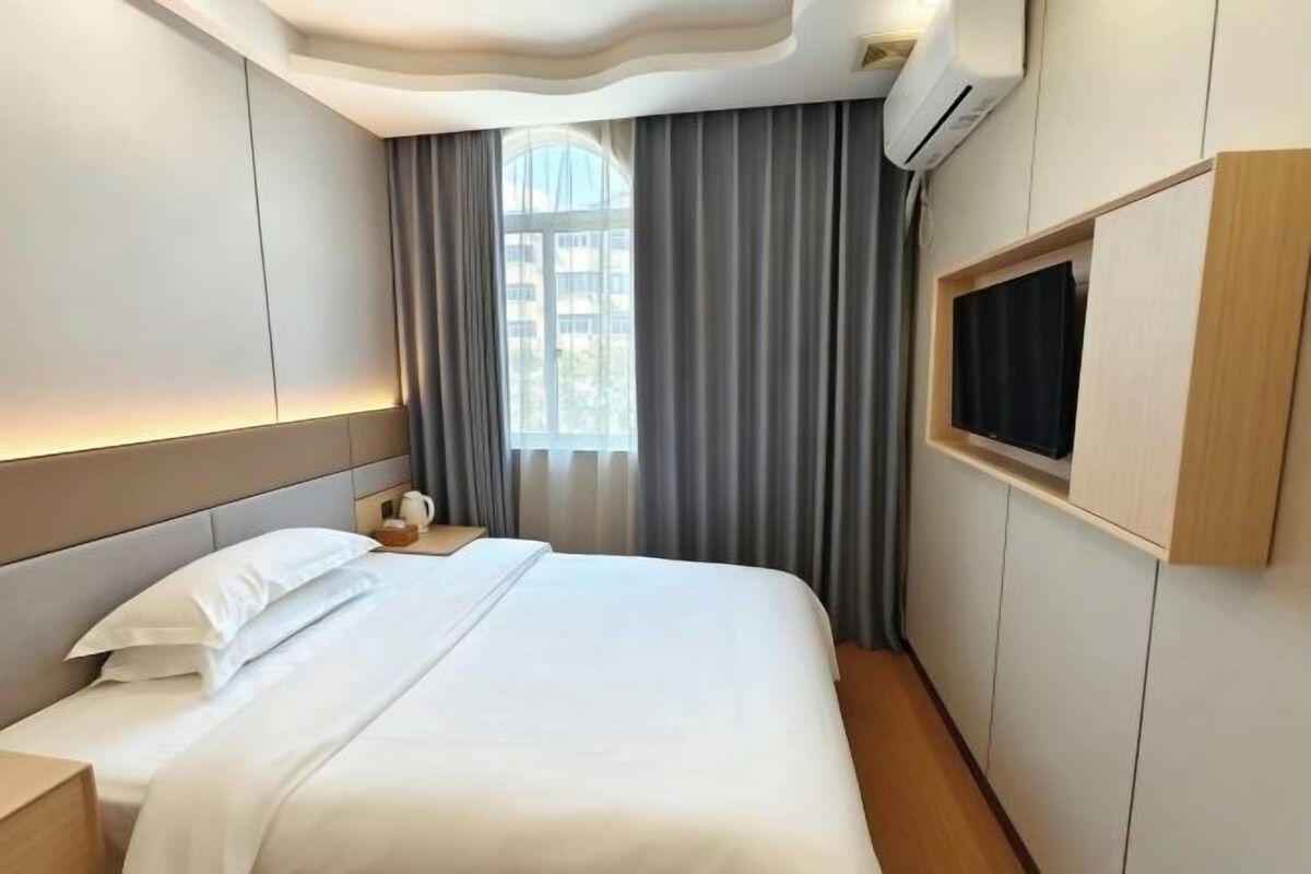 Quarto de hotel com cama, mesa de cabeceira, televisão na parede e janela com cortinas.