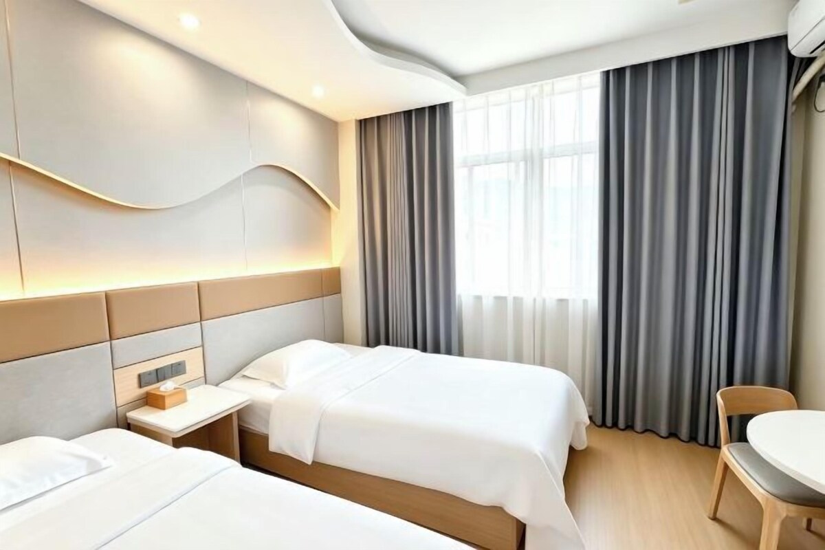 Quarto de hotel com duas camas, uma mesinha de cabeceira, uma cadeira e uma janela grande com cortinas.