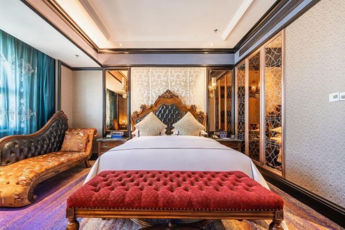 Um quarto espaçoso com uma cama grande, um banquinho estofado vermelho, um sofá e um guarda-roupa com espelho.