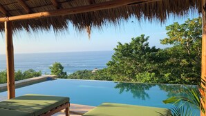 Uma piscina infinita com vista para o oceano, rodeada por vegetação exuberante e uma estrutura de telhado de palha.