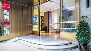 Entrada de hotel com design moderno, apresentando uma porta de vidro, um painel de sinalização digital e um vaso de planta.