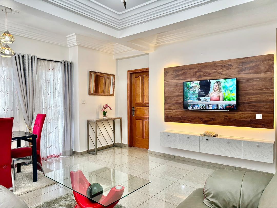 Uma sala de estar moderna com televisão de tela plana fixada em parede de madeira, mesa de centro de vidro, cadeiras vermelhas e cinzas, e uma porta de madeira.