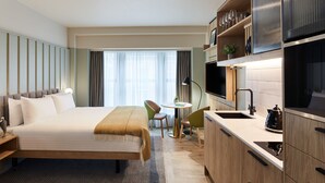 Quarto de hotel com cama, área de refeições compacta, cozinha compacta e uma janela grande com cortinas.