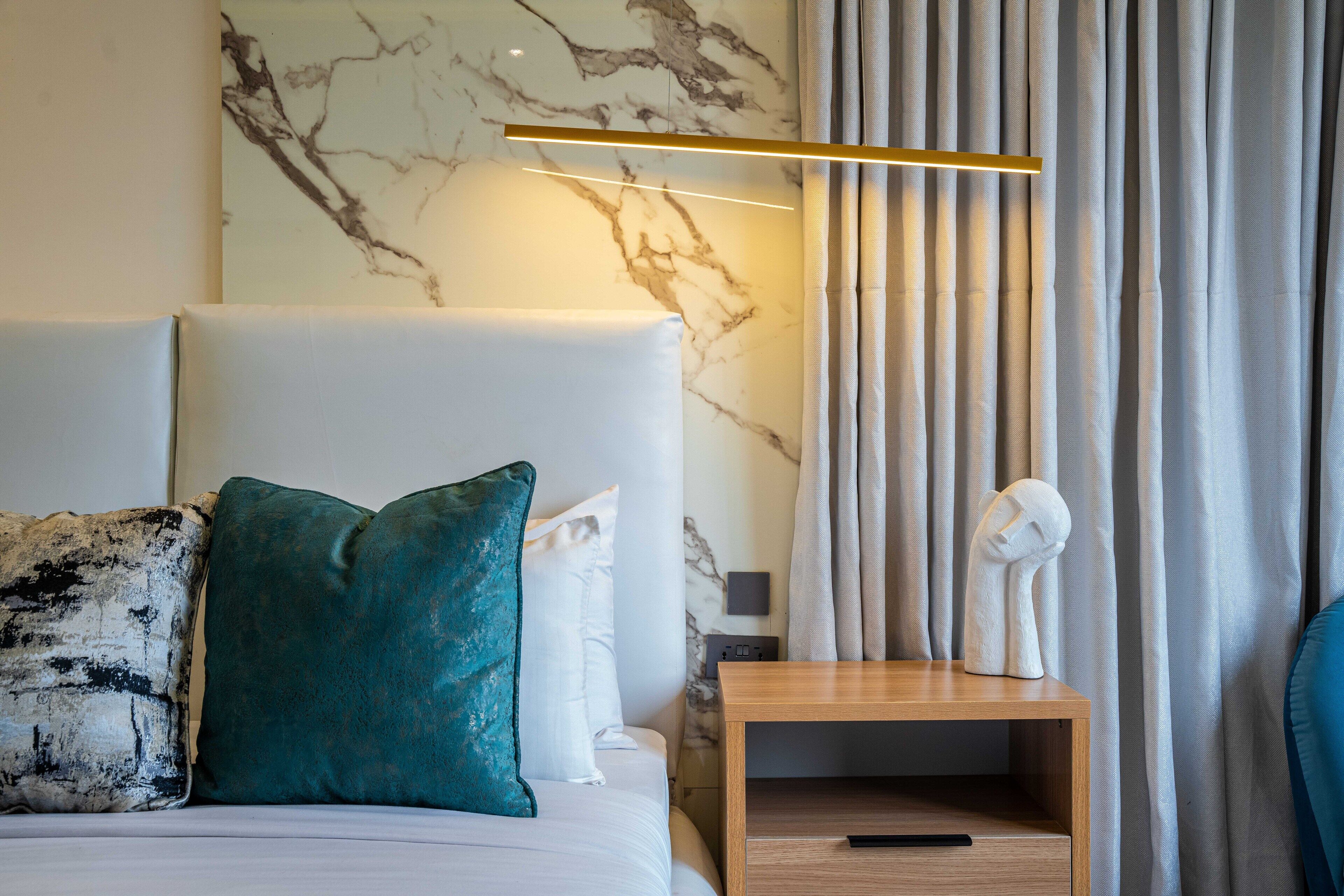 Quarto de hotel moderno com parede de mármore, cama com almofadas verde-água e brancas, mesa de cabeceira de madeira e uma escultura.