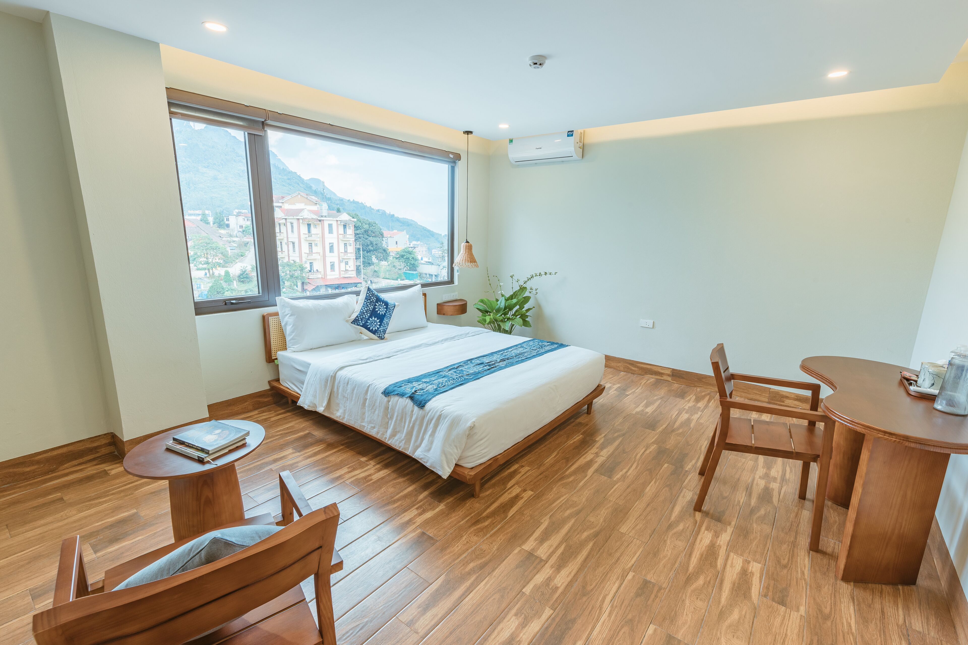 Quarto de hotel com uma cama grande, piso de madeira, uma pequena mesa, uma cadeira, uma escrivaninha e vista para montanhas e edifícios.