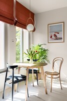 Uma área de refeições com uma mesa redonda, duas cadeiras, uma planta em vaso e um lustre.