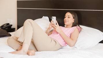 Uma pessoa deitada em uma cama, com um smartphone, um telefone de mesa e um travesseiro.