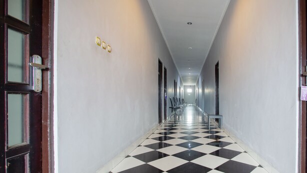 Corredor de hotel com piso xadrez, portas em ambos os lados e uma fileira de cadeiras ao fundo.