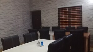 Uma sala de conferências com uma mesa branca, cadeiras pretas, um papel de parede estampado, uma janela com persianas e uma porta.