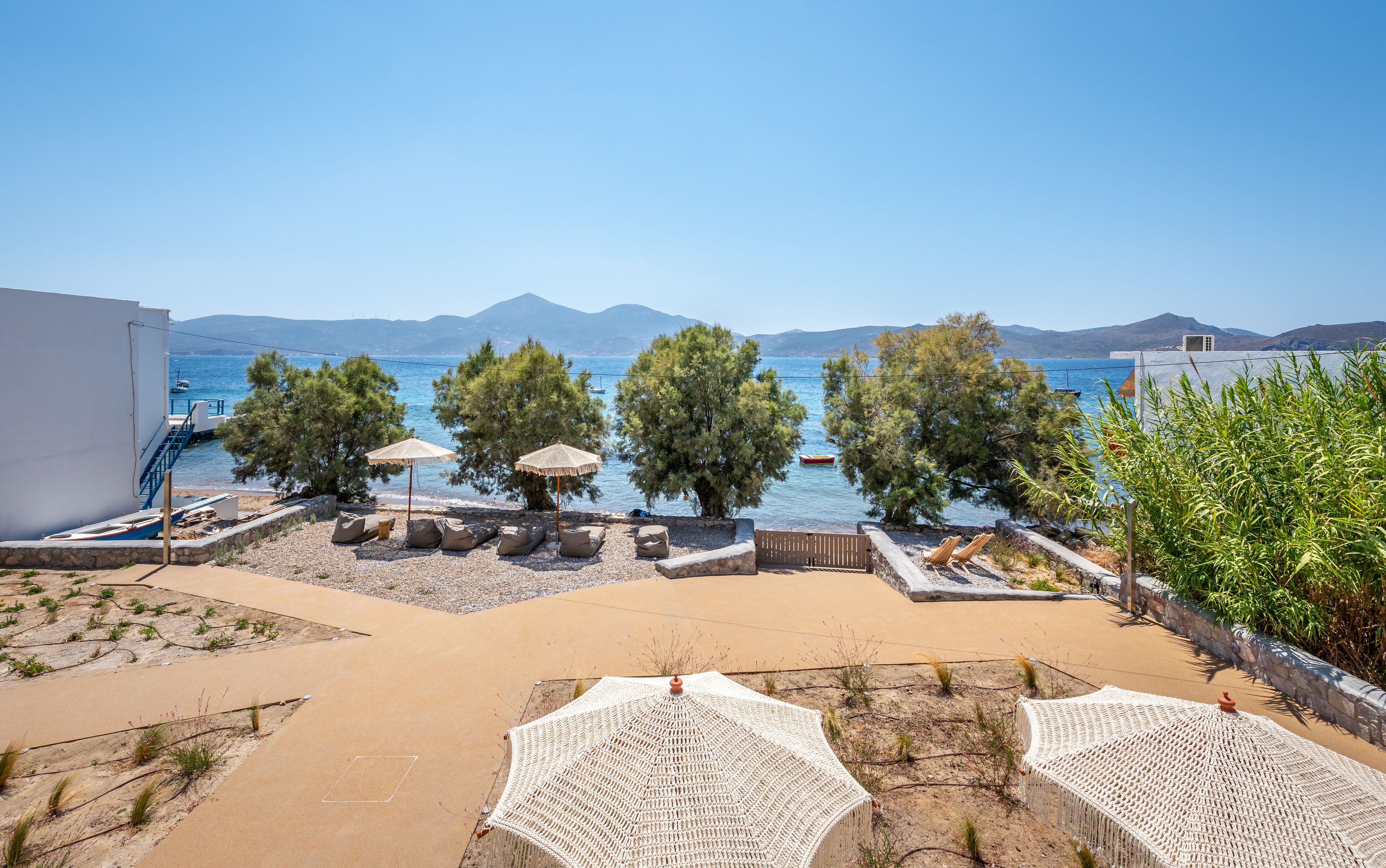 Um terraço à beira-mar com guarda-sóis, mesas e cadeiras, e vista para o mar e montanhas.
