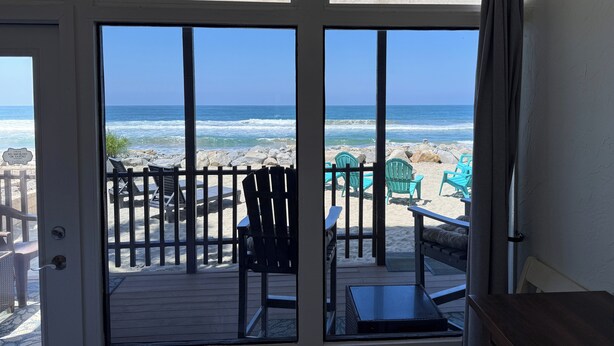 Vista de um quarto com uma grande janela para uma praia, com cadeiras e um deck de madeira.