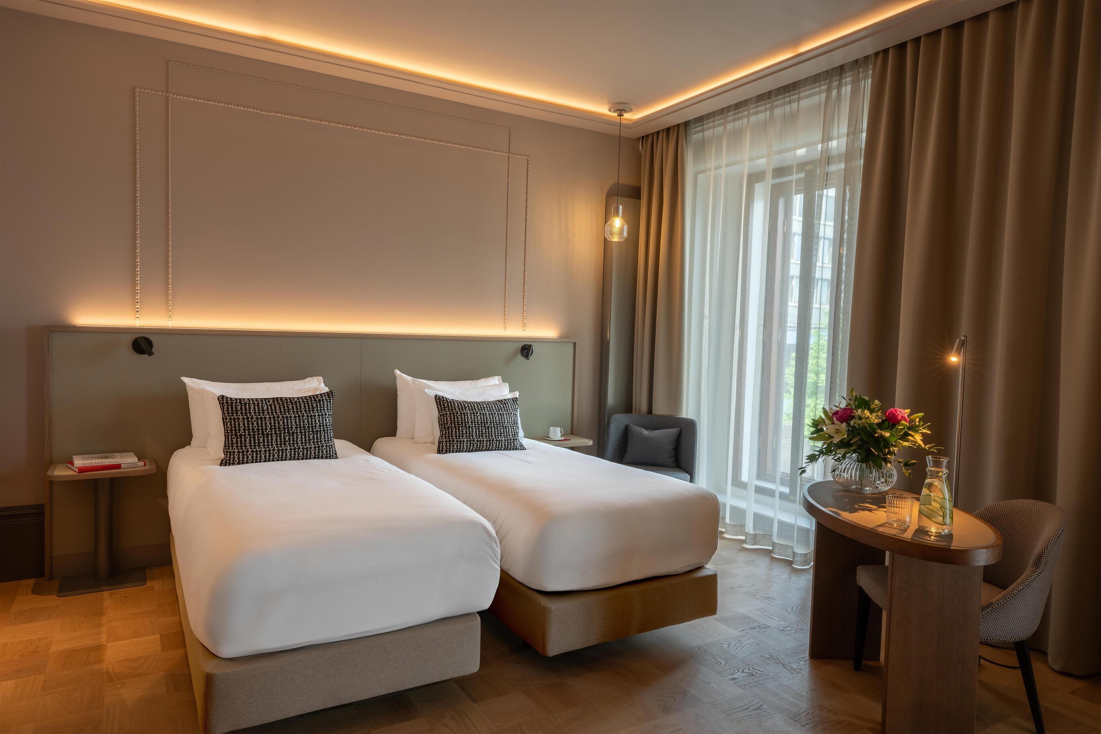Quarto de hotel com duas camas, uma mesinha com um vaso de flores e uma cadeira.