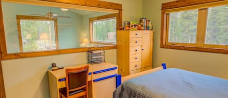 Quarto com cama, escrivaninha com cadeira, guarda-roupa e duas janelas com vista para árvores.