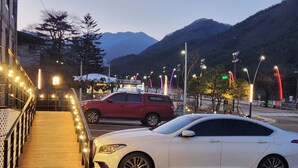 Um carro branco estacionado na rua, com um SUV vermelho ao fundo, uma ponte de madeira iluminada e montanhas à distância.