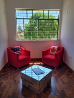 Um quarto com duas poltronas vermelhas, uma mesa de centro de vidro e uma janela grande com vista para árvores e edifícios.