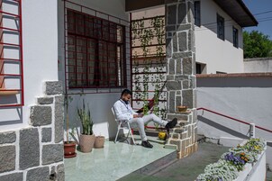 Um homem sentado numa cadeira em uma escadaria de pedra em frente a um edifício com guarda-corpo vermelho, plantas em vasos e uma janela com padrão de grade vermelha.
