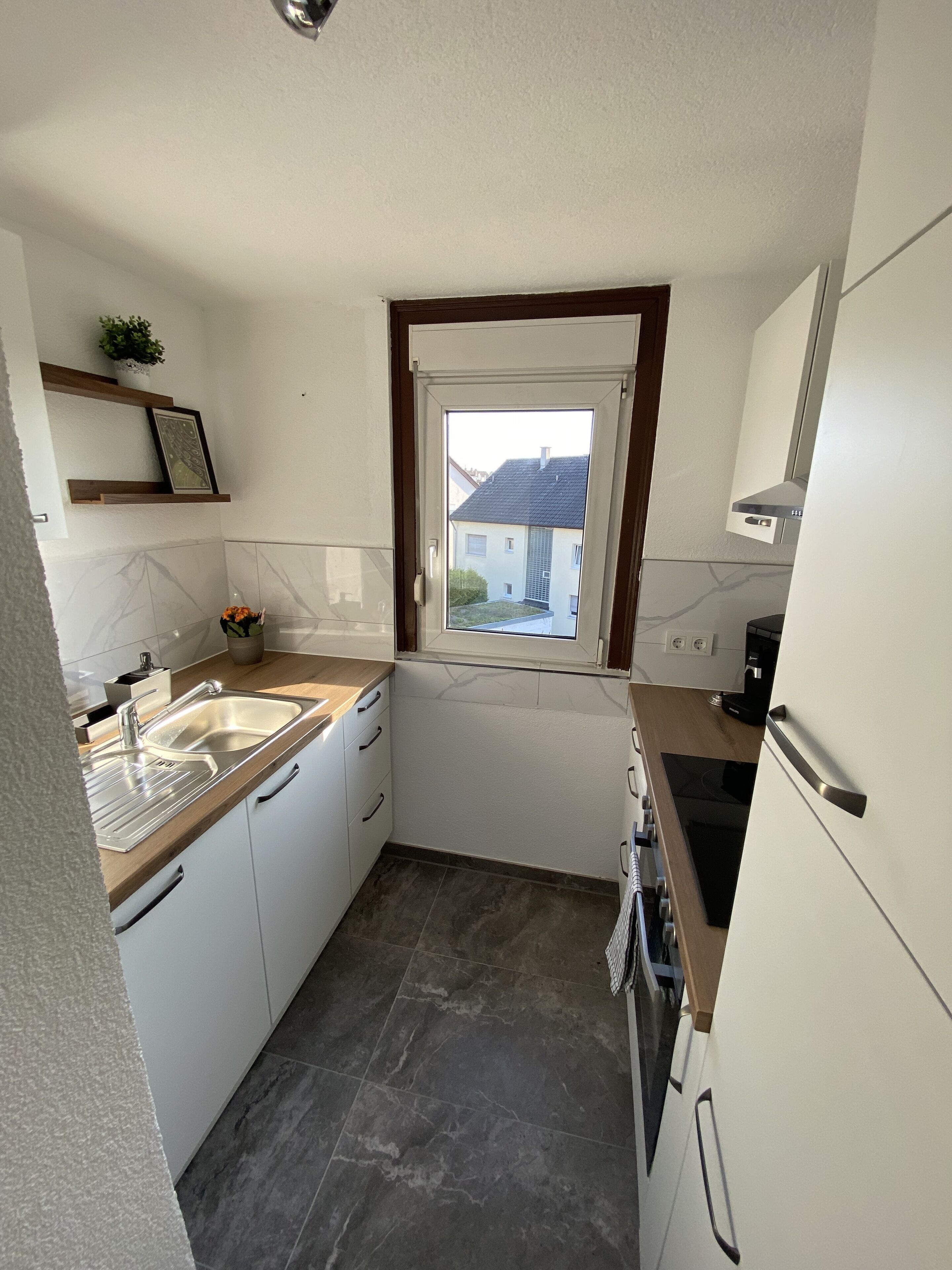 Uma cozinha compacta com armários brancos, uma pia de aço inoxidável, uma janela com vista para uma casa e um forno embutido.