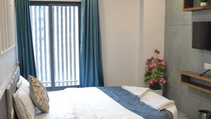 Quarto de hotel moderno com cama curva, secretária pequena, televisão e janela com cortinas azuis.